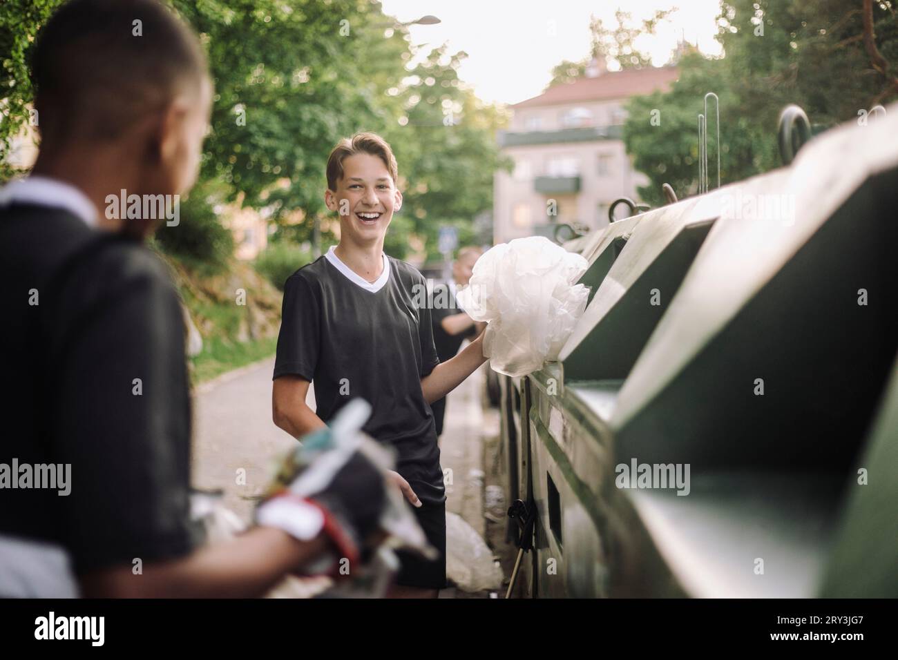 Happy teenage boy putting plastic garbage bag in recycling bin near ...
