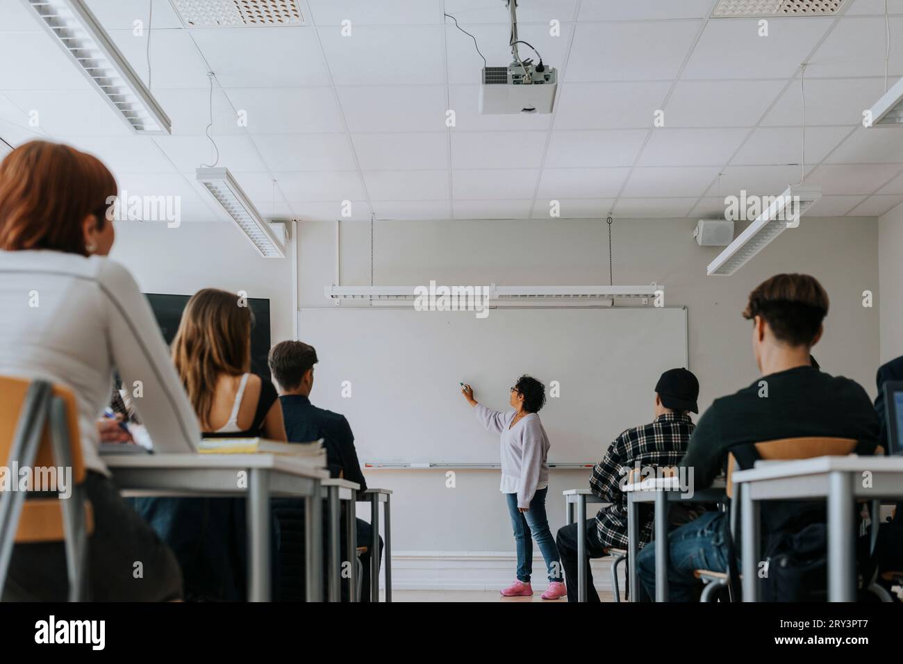 Students sitting at desk while teacher writing on whiteboard in ...