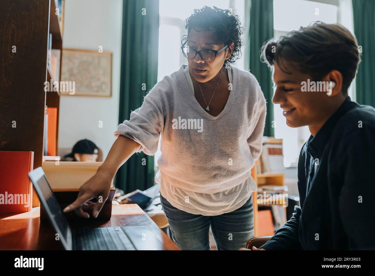 Teacher pointing at laptop while teaching smiling male student in ...