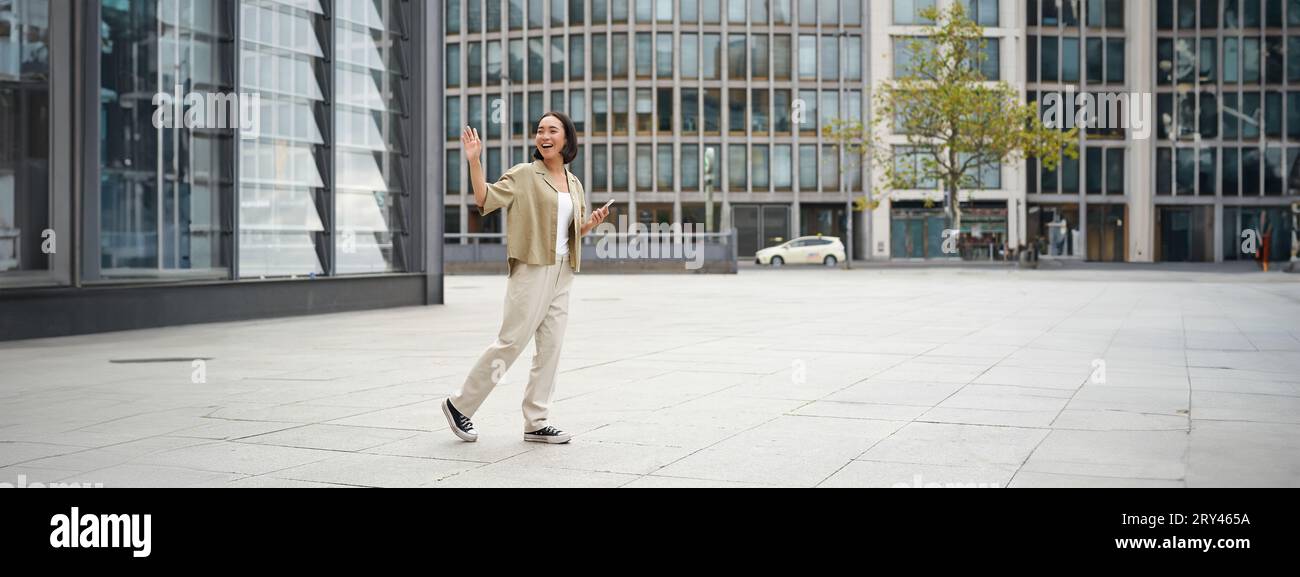 Happy asian girl saying bye to someone, waving at friend and walking on ...