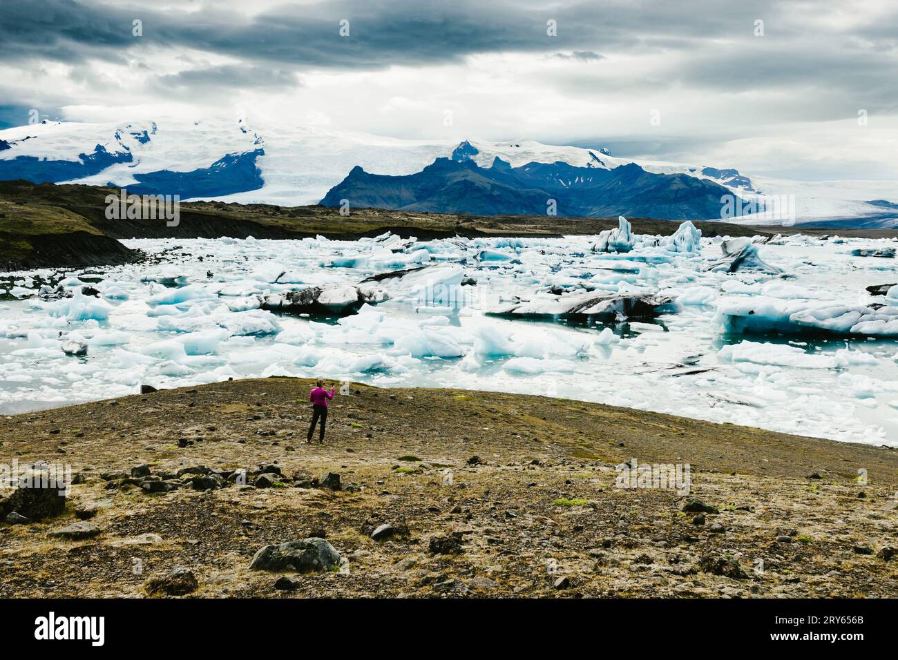 Woman hikes along glacial lagoon with a glacier on mountain behind ...