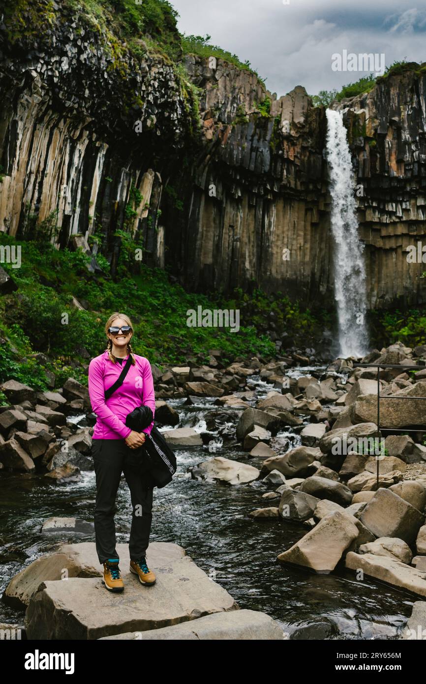 Woman smiles in front of waterfall over basalt cliff with greenery ...