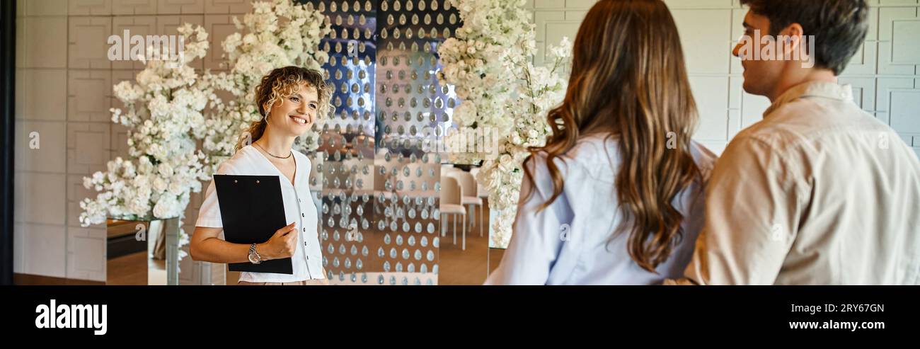 event organizer with clipboard smiling near couple in banquet hall with ...