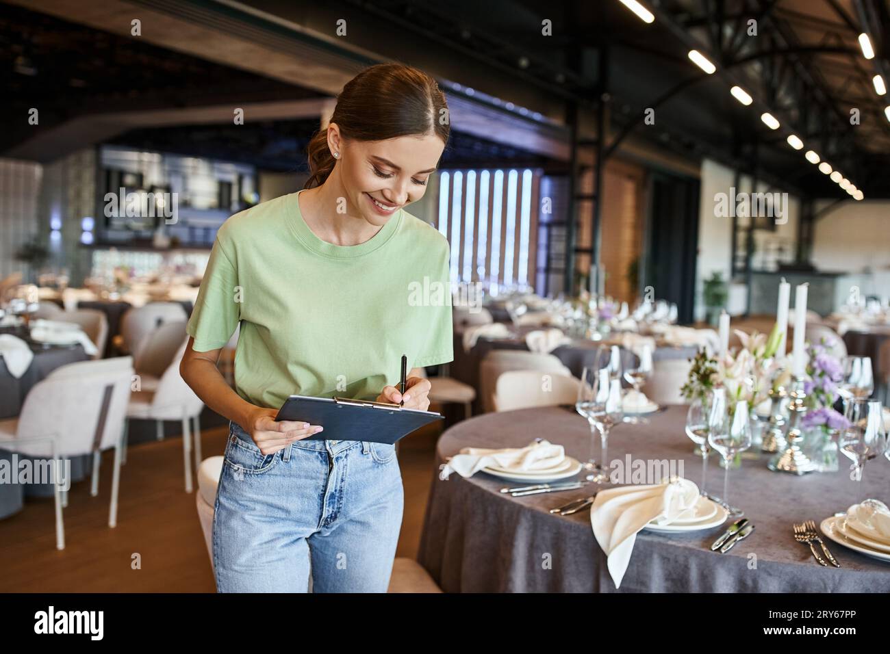 joyful event manager writing notes on clipboard near tables with ...