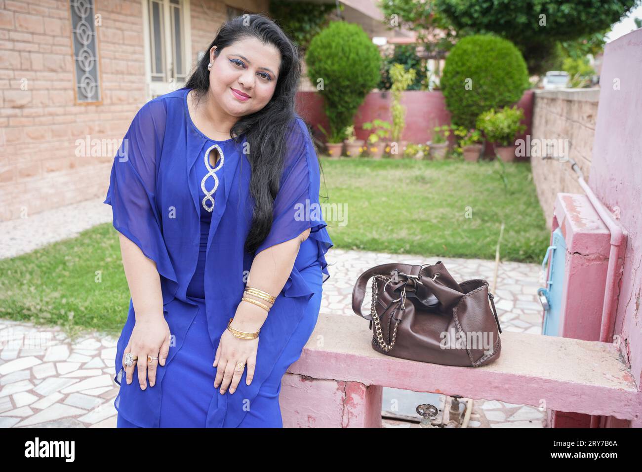 Happy young indian plus size woman wearing blue dress sitting outside ...