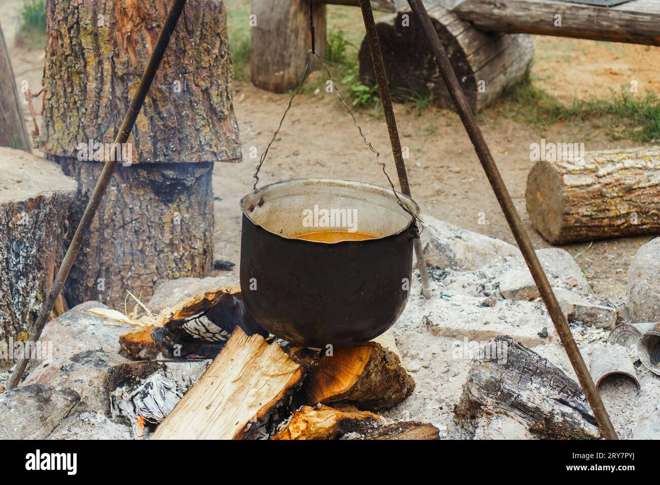 Metal vessel with liquid meal inside hanging on tripod over firewood ...
