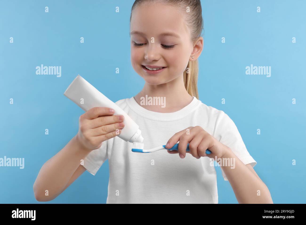 Happy girl squeezing toothpaste from tube onto toothbrush on light blue ...