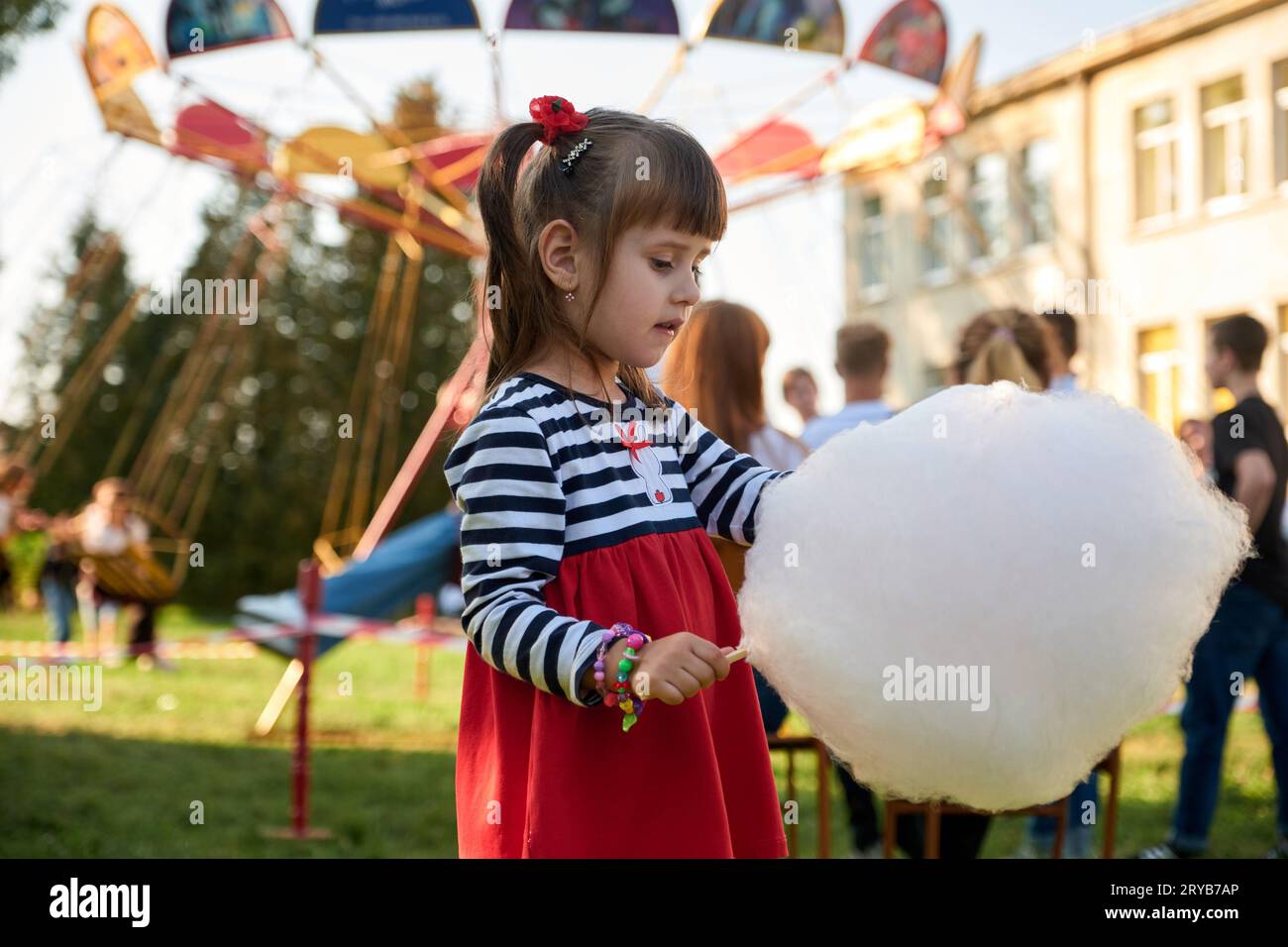 Cute little girl eating sweet cotton wool against the backdrop of rides ...