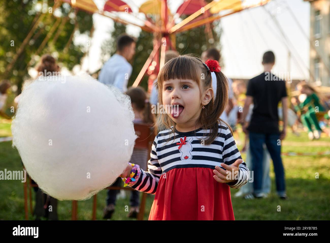 Cute little girl eating sweet cotton wool against the backdrop of rides ...