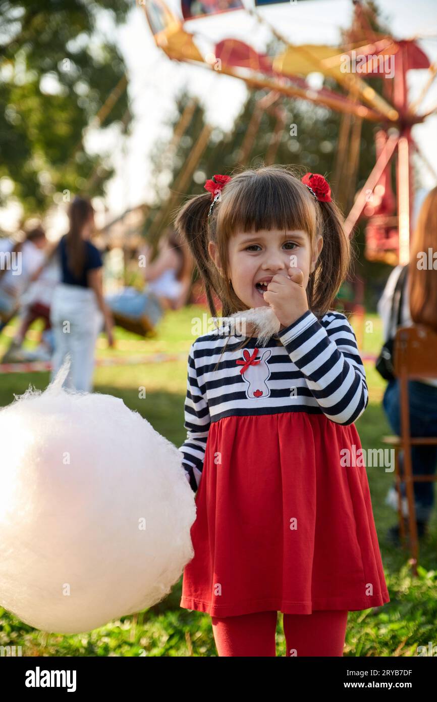 Cute little girl eating sweet cotton wool against the backdrop of rides ...
