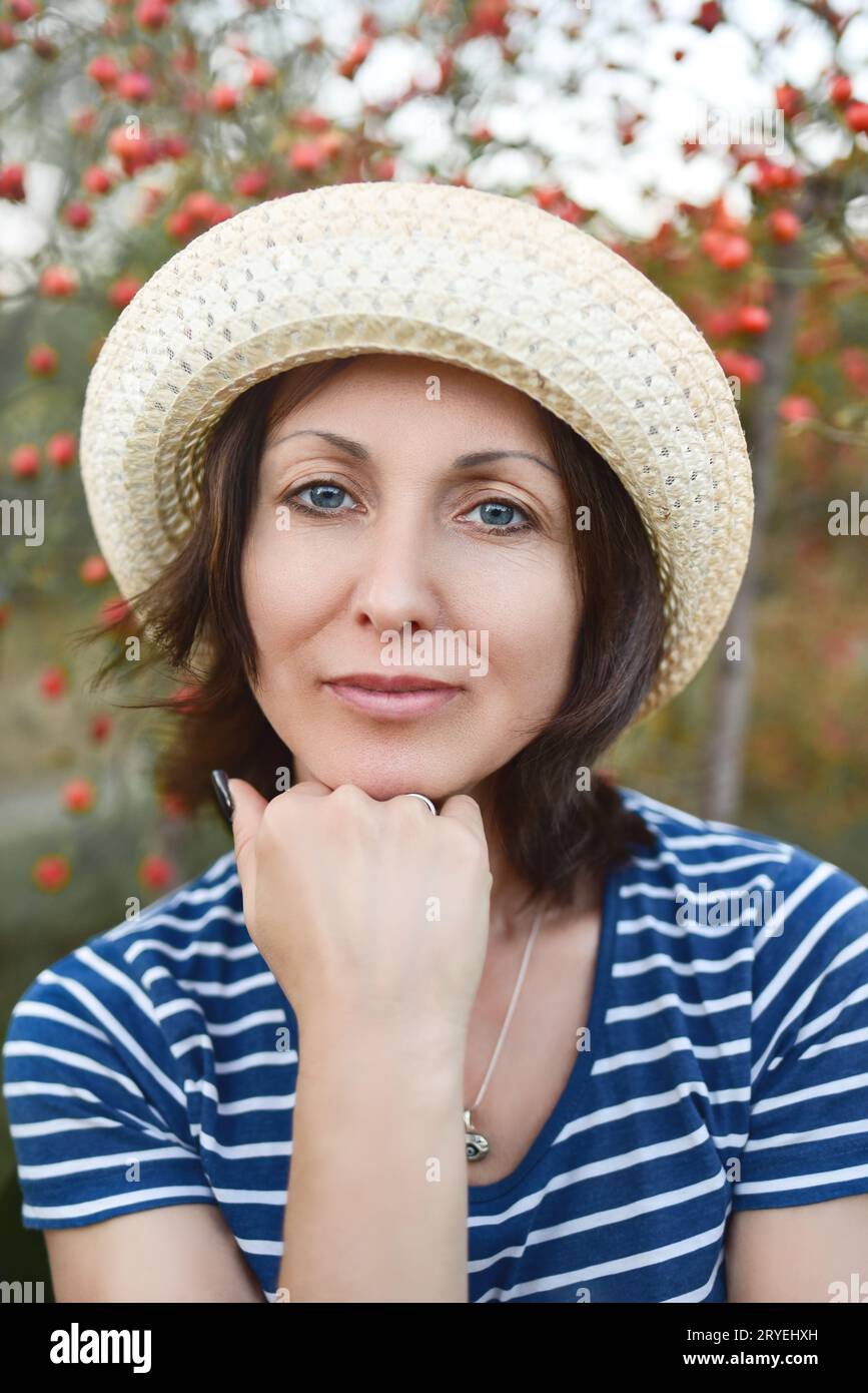 Image of beautiful middle-aged woman wearing straw hat laughing and looking at camera while ...