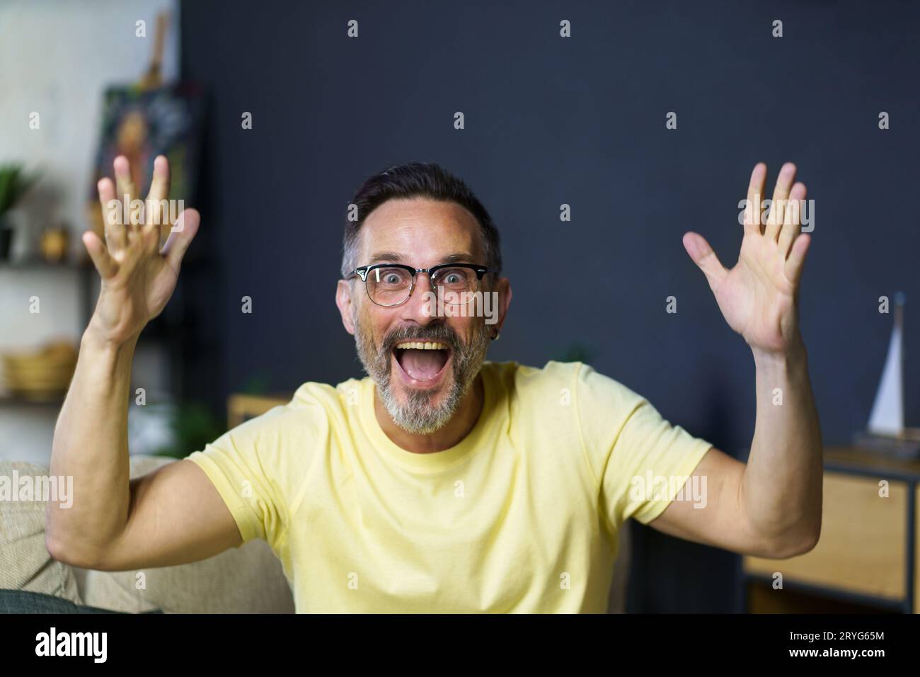 Happy excited middle aged grey haired man sitting on the sofa with hands up in the air looking ...