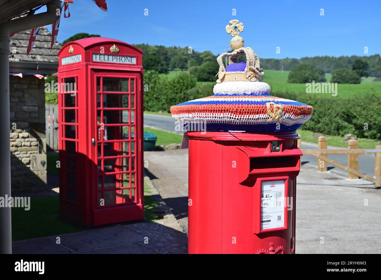 A free-standing letter box with a knitted top, beside a red telephone ...