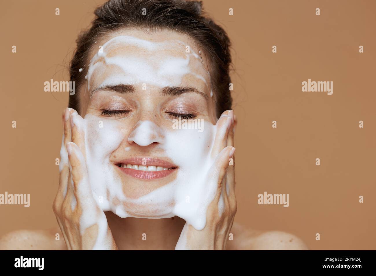 relaxed young woman with foaming face wash washing face against beige ...