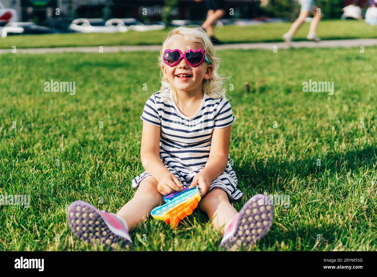 A little adorable smiling three-year-old girl is sitting on the lawn ...