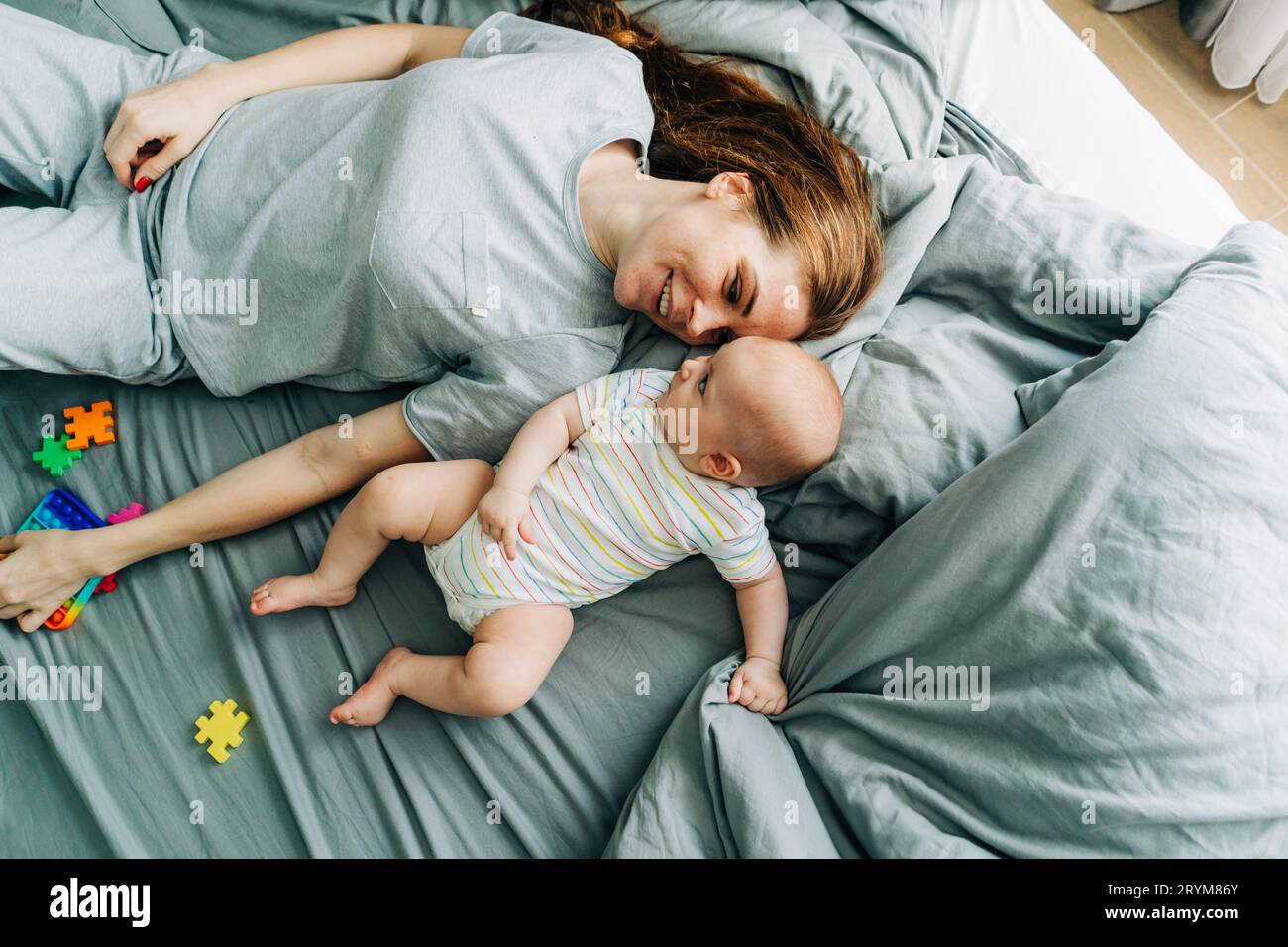 Adorable young red-haired mom with her baby daughter lie in gray bed ...