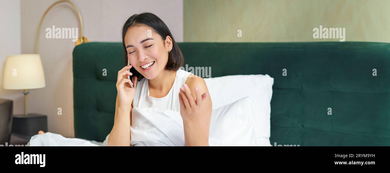 Close up portrait of cute asian girl in bed, talking on mobile phone ...