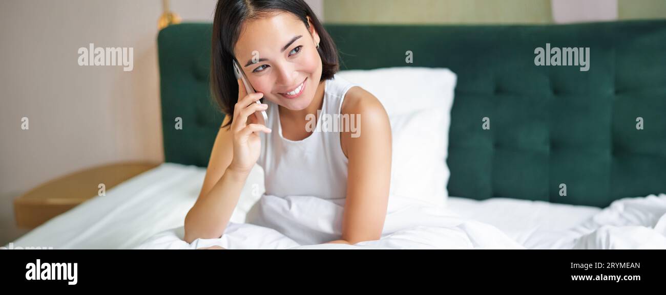Smiling korean girl talks on mobile phone and lying in bed. Cute woman ...
