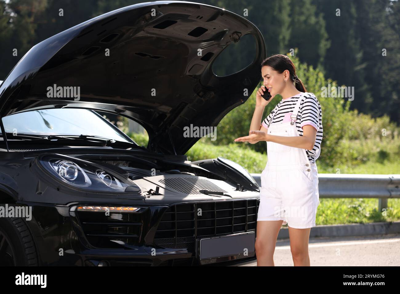 Stressed woman talking on smartphone while looking under hood of broken car outdoors Stock Photo ...