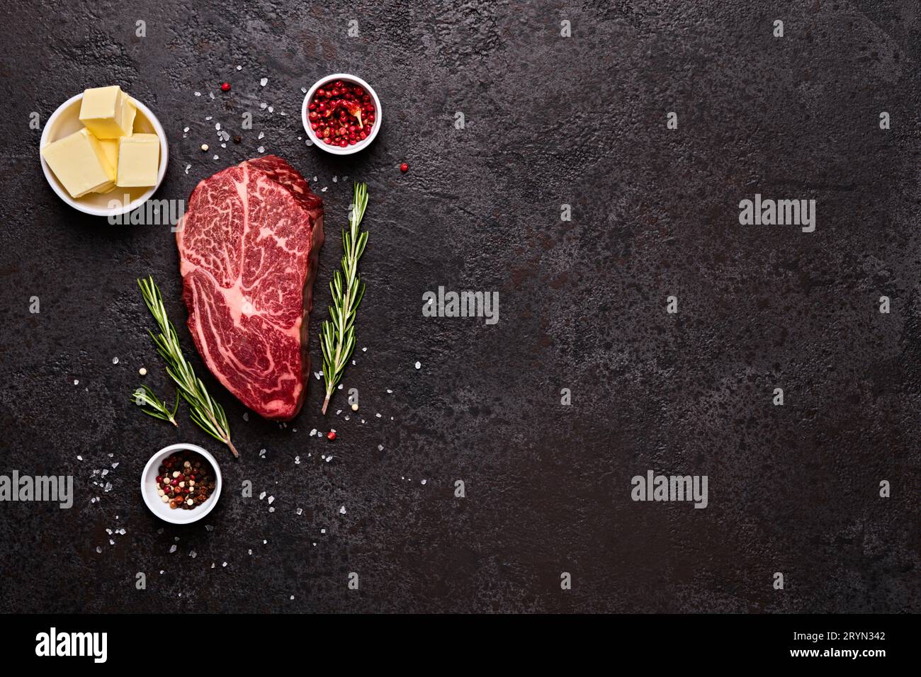 Fresh raw marbled beef rib eye steak, butter and spices on black stone ...