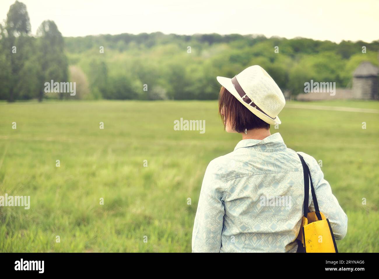 Back view of brunette woman wearing shirt , straw hat and bag walks on ...