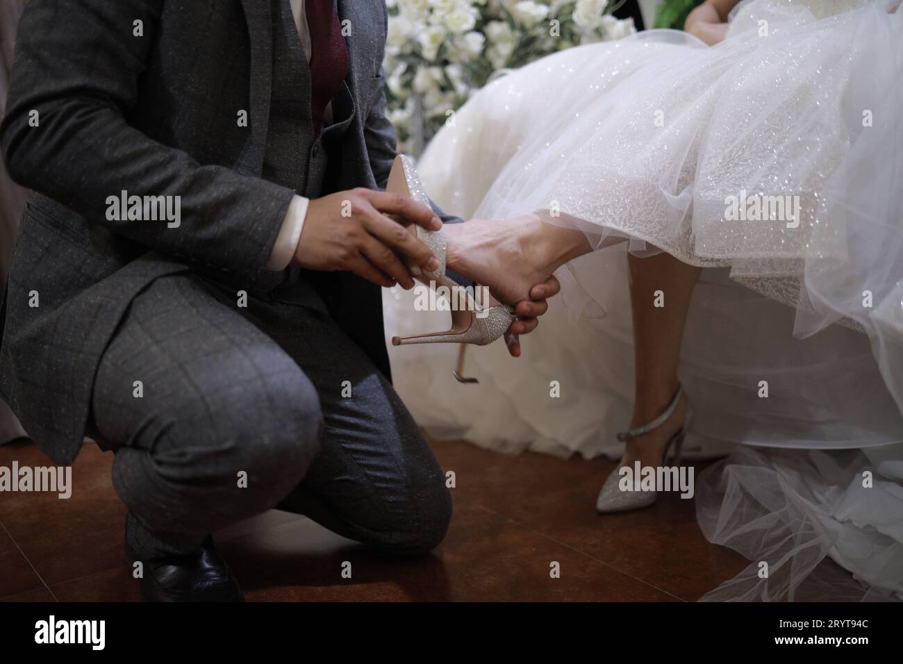 A groom in formal attire kneels while cutting a three-tiered white ...