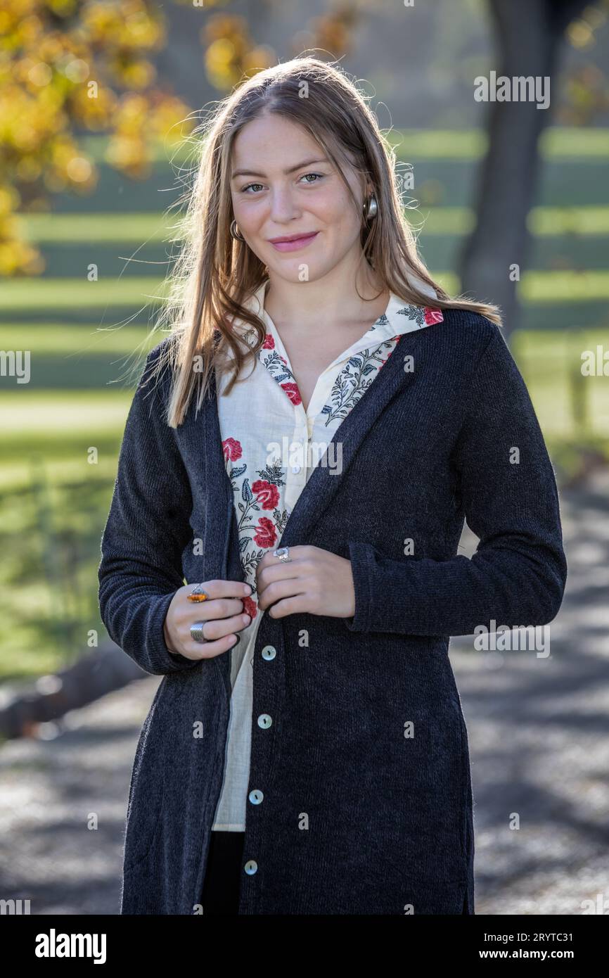 Beneath the autumn trees, a young woman buttons up her cardigan as she ...