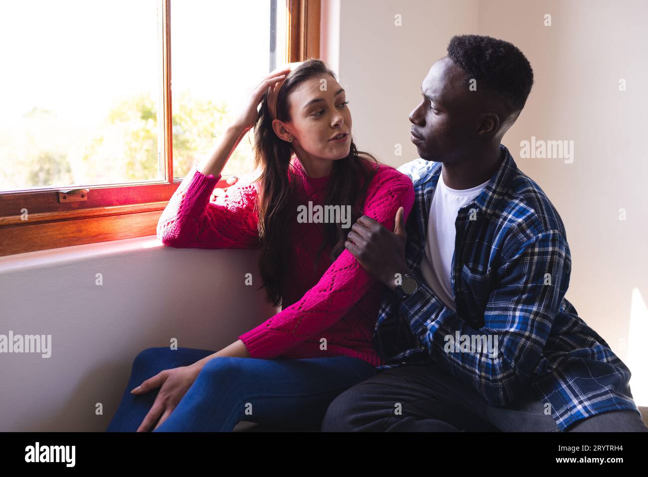 Diverse couple embracing and talking next to window at home Stock Photo ...