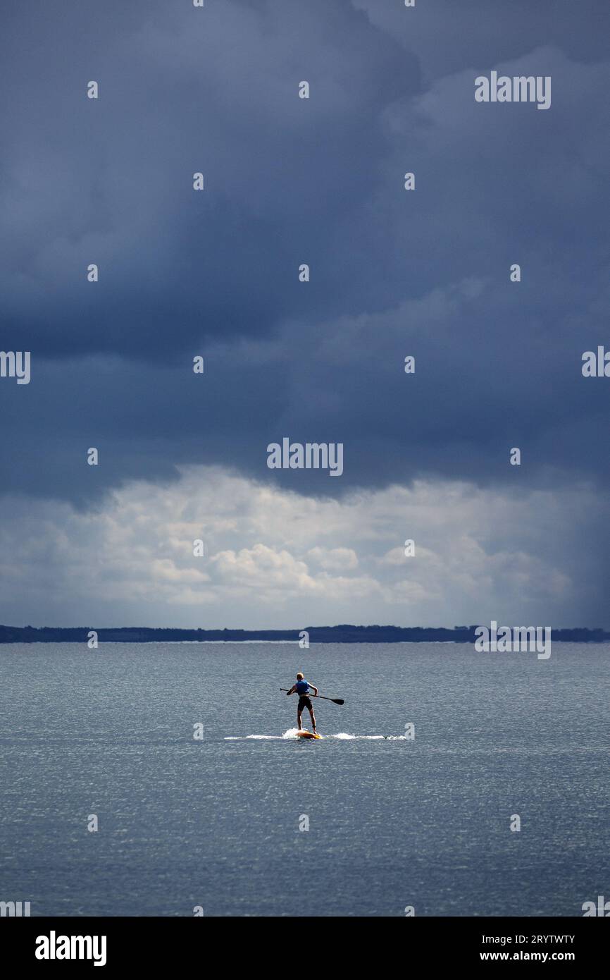 A young adult is seen surfboarding through choppy waters as a grey sky ...