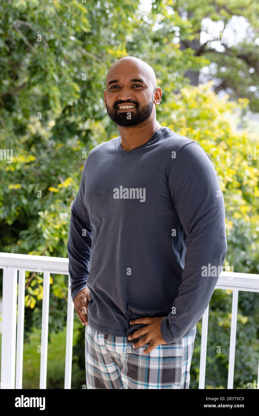 Portrait of happy biracial man in pyjamas standing on sunny balcony at home in front of treetops ...