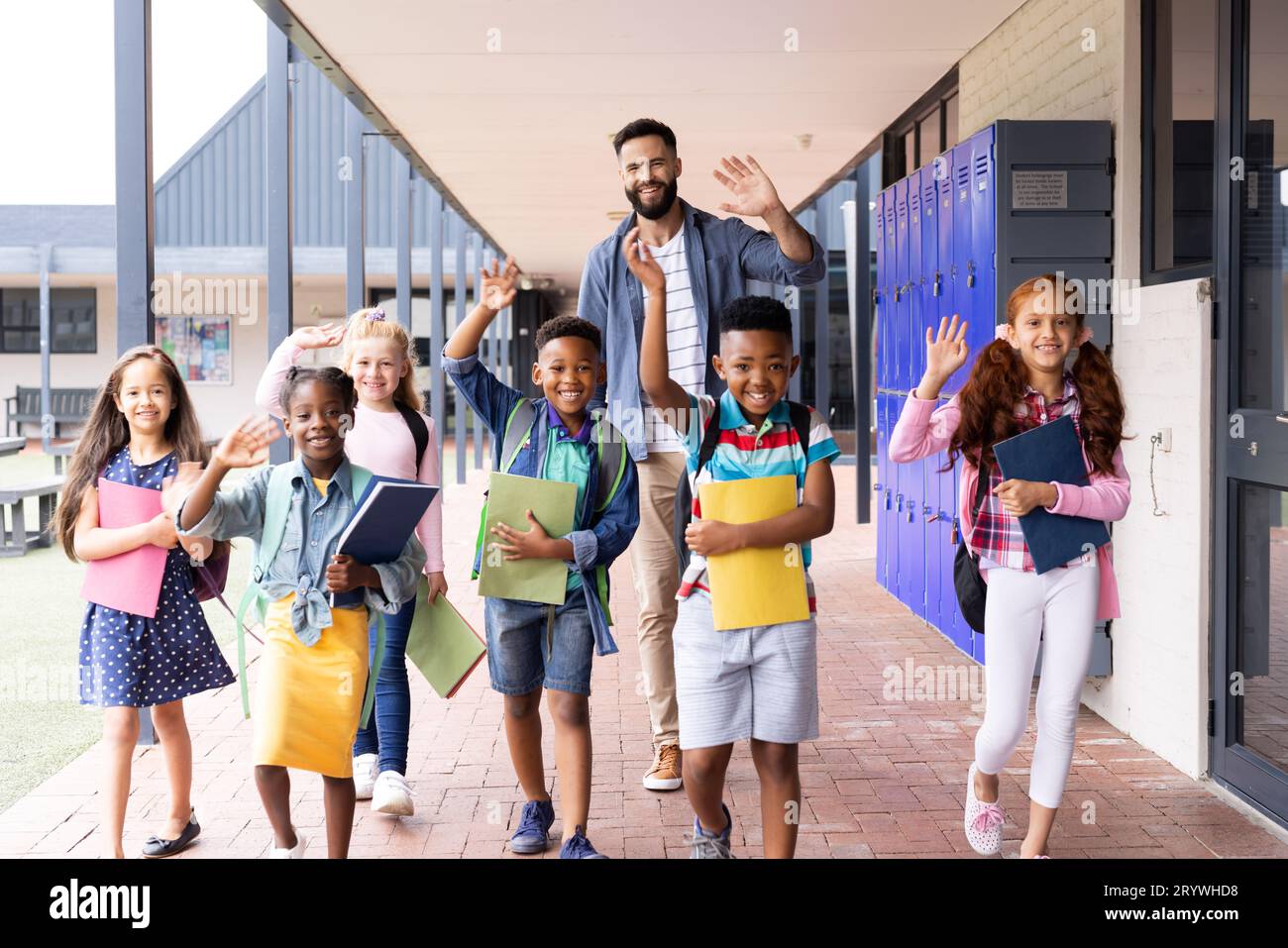 Diverse, happy male teacher and elementary schoolchildren waving in school corridor, copy space ...