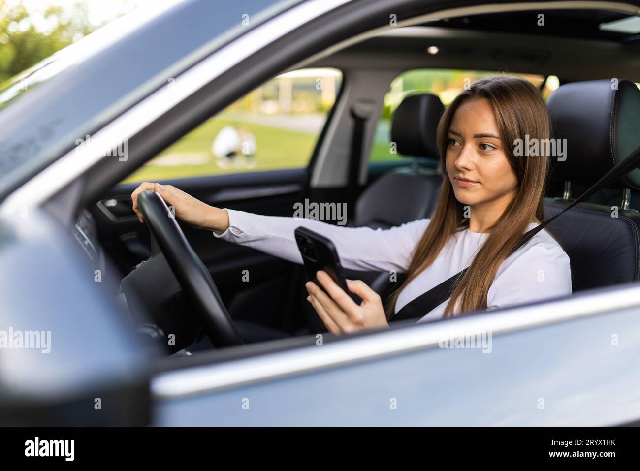 Cheerful young woman sitting in a car in the drivers seat looking into a smartphone, paying for ...