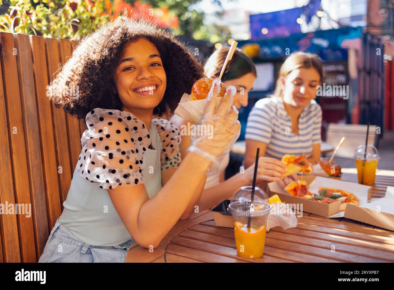 Cute smiling teenage girls are sitting in open air cafe and eating fast ...