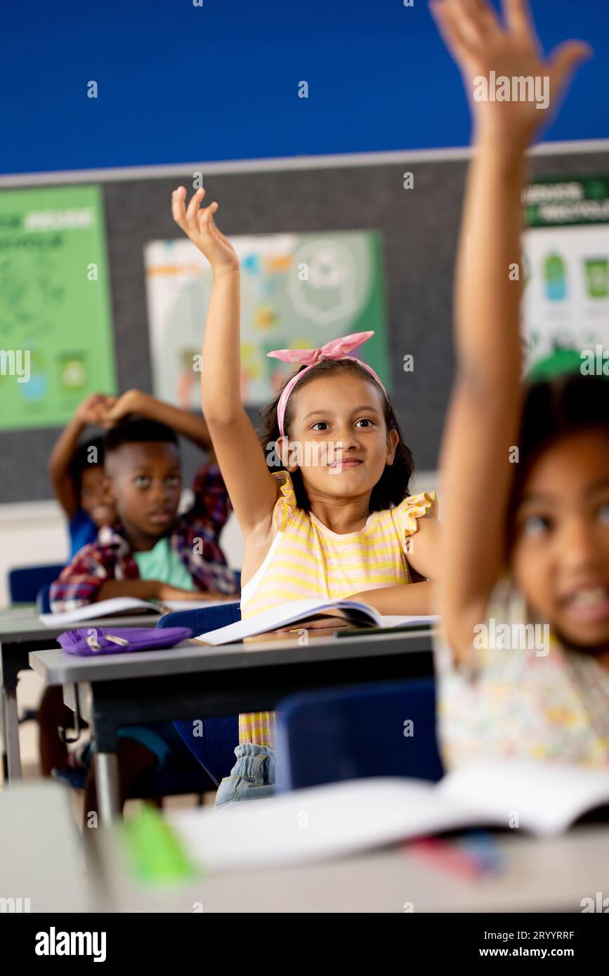 Diverse elementary schoolchildren sitting at desks and raising hands in ...