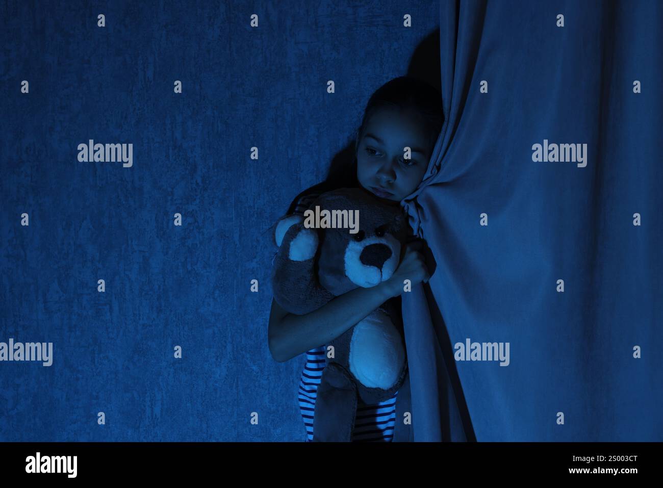 Scared girl hiding with teddy bear behind curtain at night. Space for ...