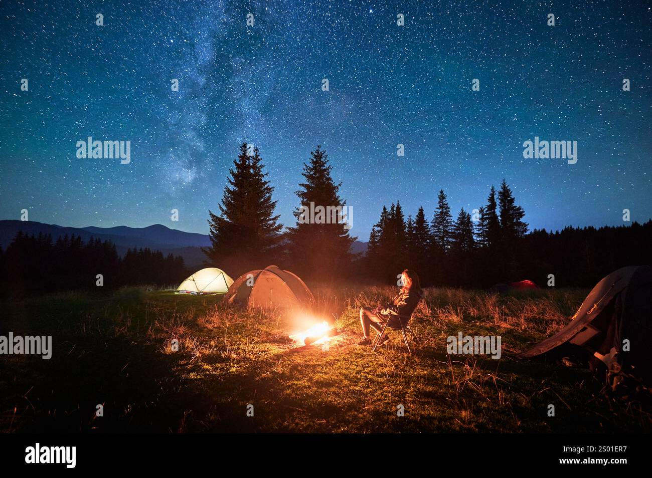 Hiker woman relaxes by campfire, illuminated by its warm glow, with ...