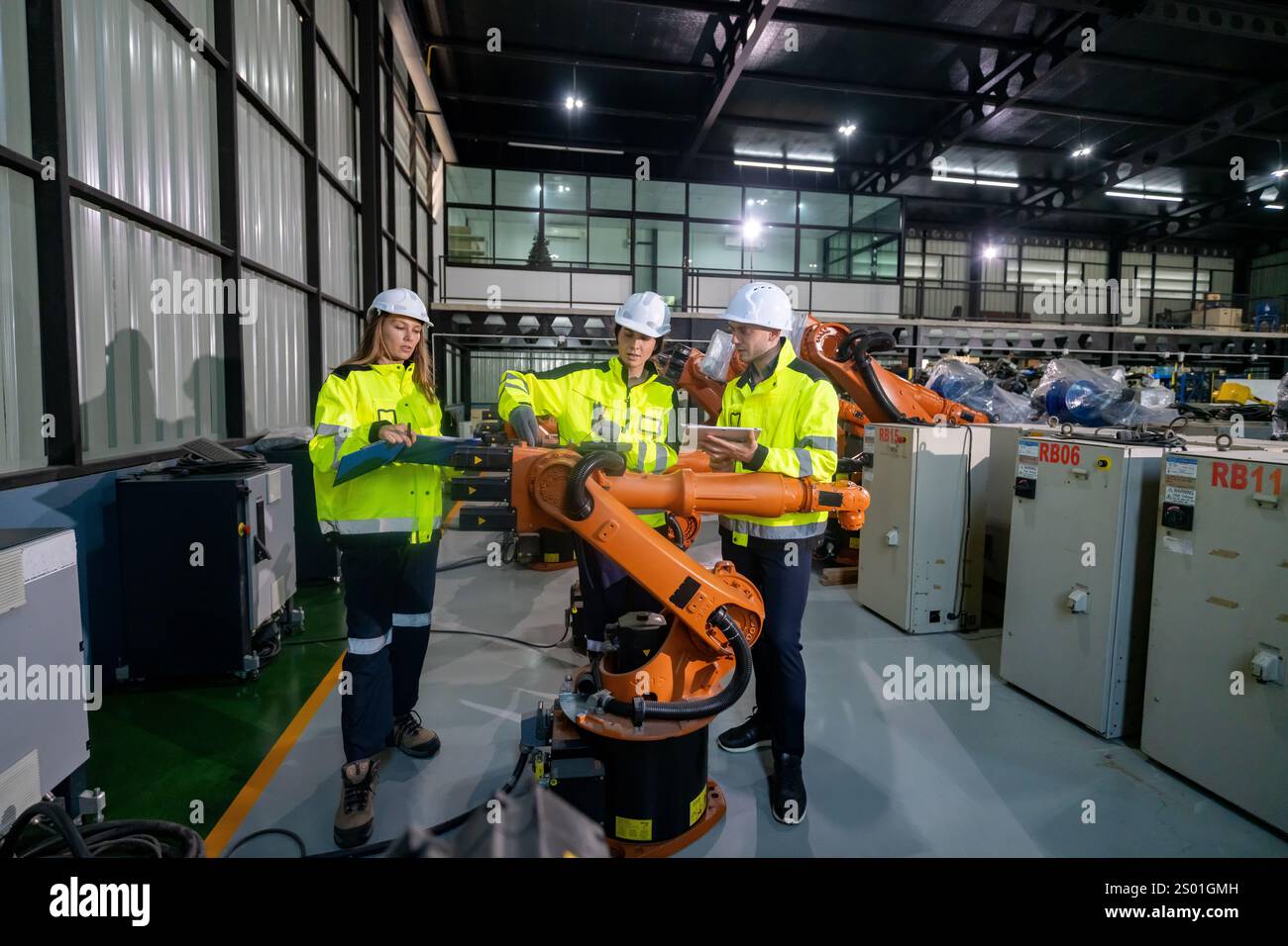 Three engineers in safety gear examine and interact with a robotic arm ...