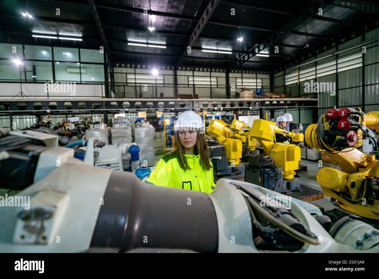 A woman in a fluorescent work jacket and hard hat inspects robotic arms ...