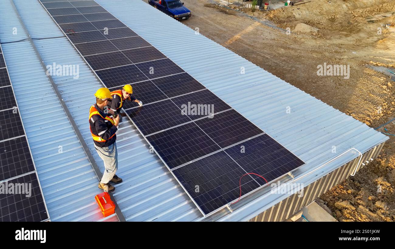 Two workers are on a rooftop attaching solar panels while wearing safety helmets. The setting is ...