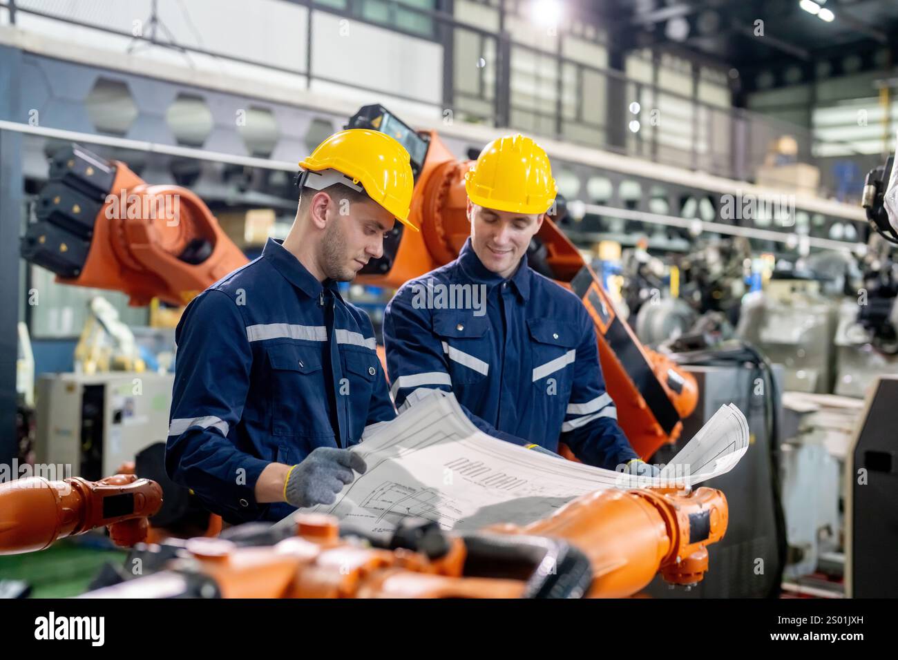 Two engineers in yellow safety helmets examine blueprints near robotic ...