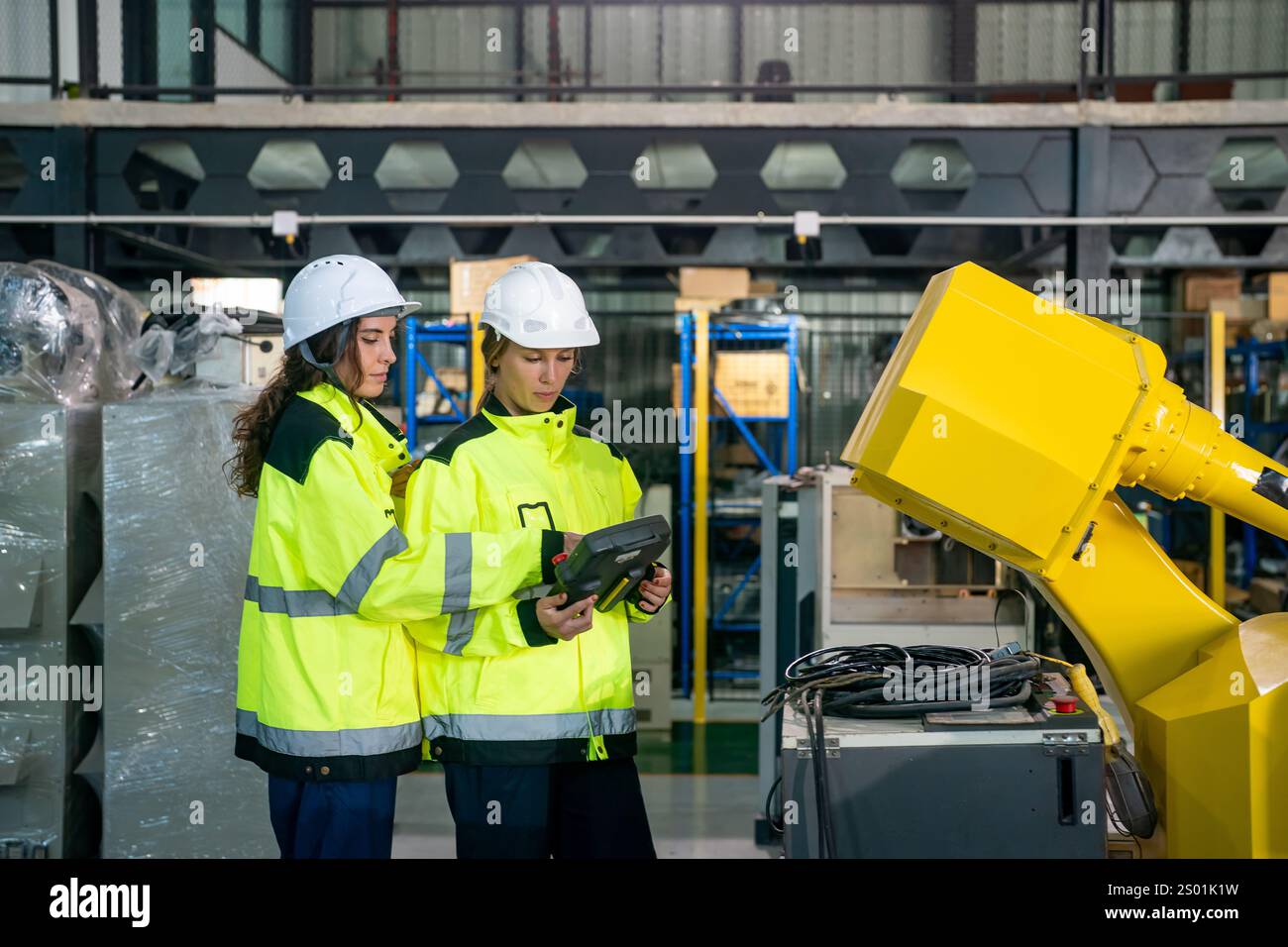 Two workers wearing safety gear are examining robotic equipment in a well-lit industrial ...