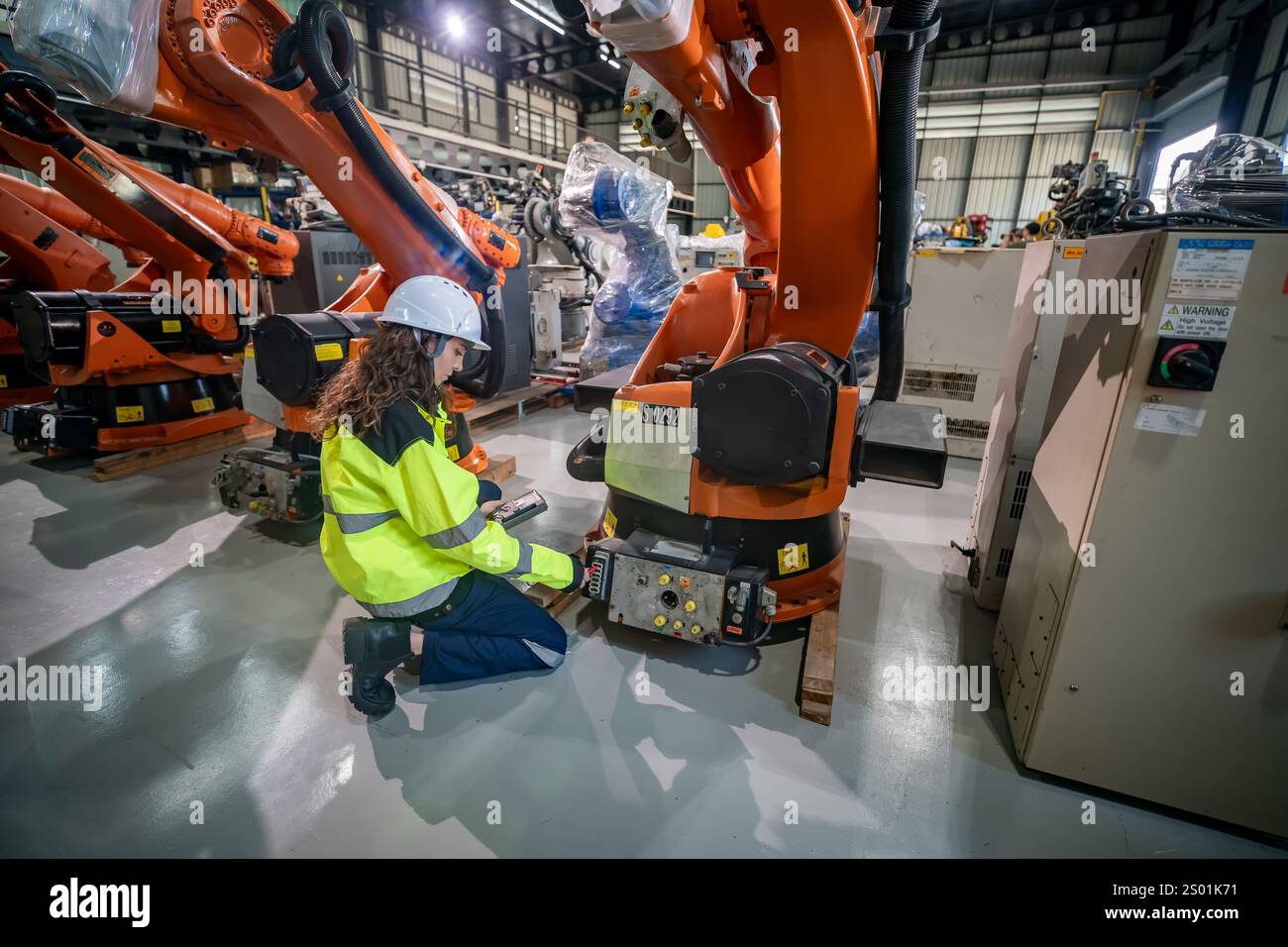 A technician wearing a hard hat and safety gear examines a robotic arm ...