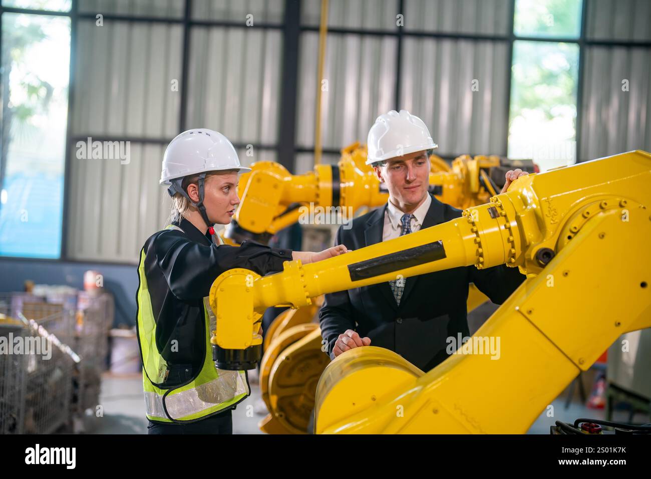 An engineer and an executive collaborate in a robotics factory, examining a robotic arm closely ...