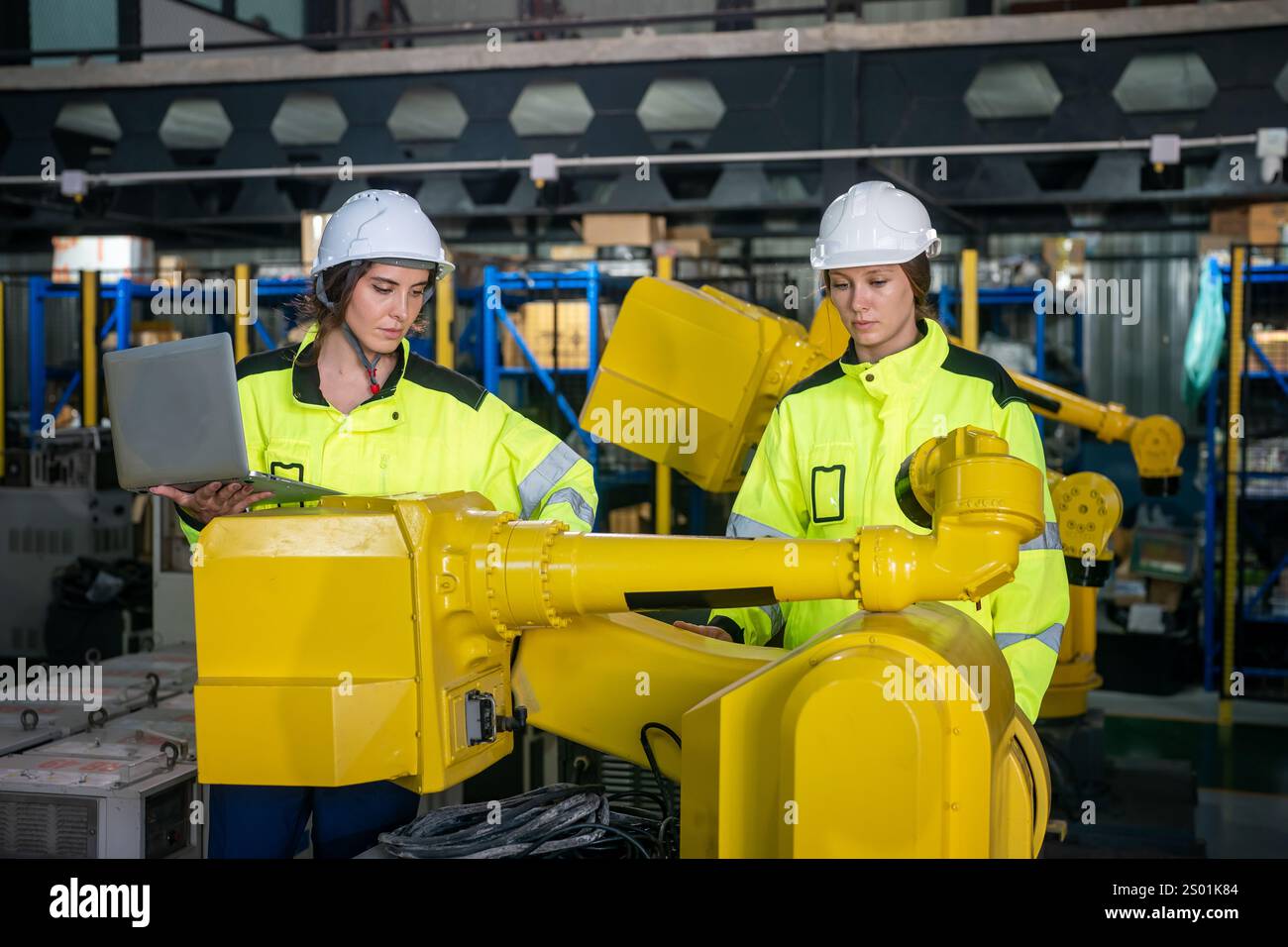 Two engineers in safety gear monitor yellow robotic arms in a modern manufacturing facility ...