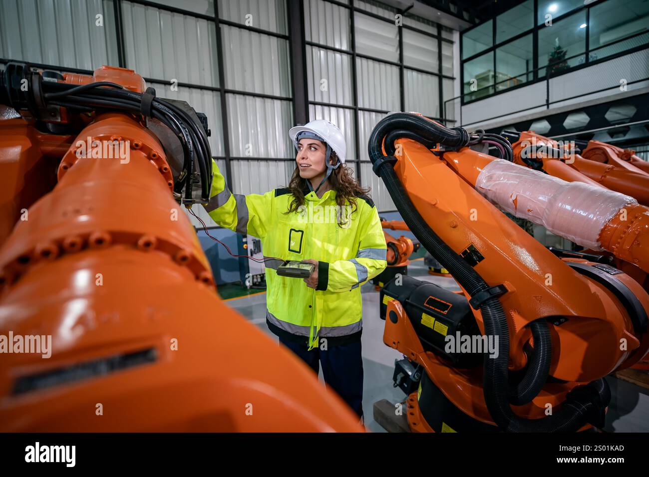 A woman in a reflective safety jacket examines robotic arms in a modern ...