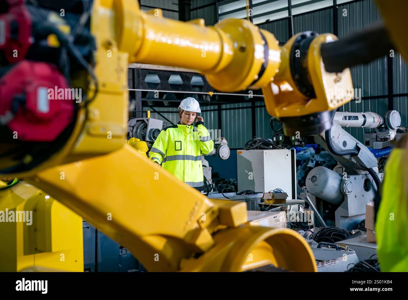 Equipment and robots are visible in a workshop while a worker in safety gear observes operations ...