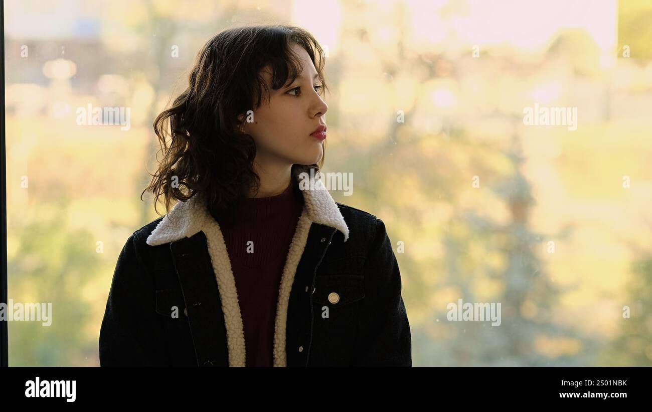 A contemplative girl looks outside through a window, bathed in soft ...