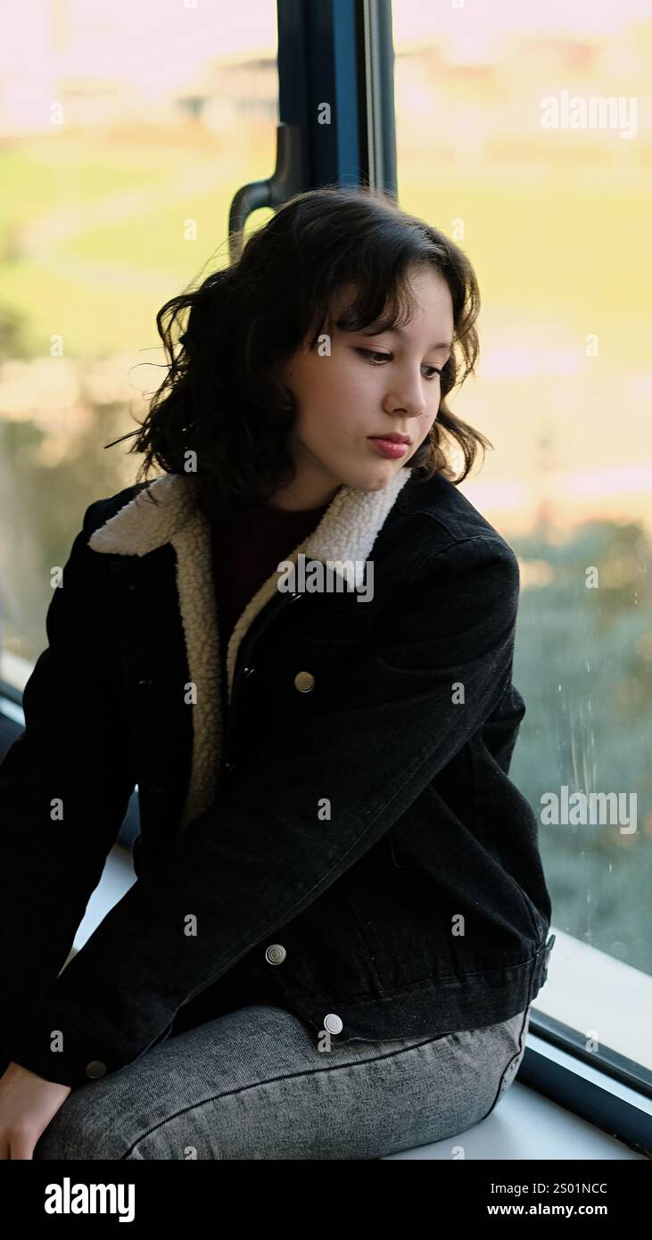 A girl leans against a windowsill, her expression calm and ...