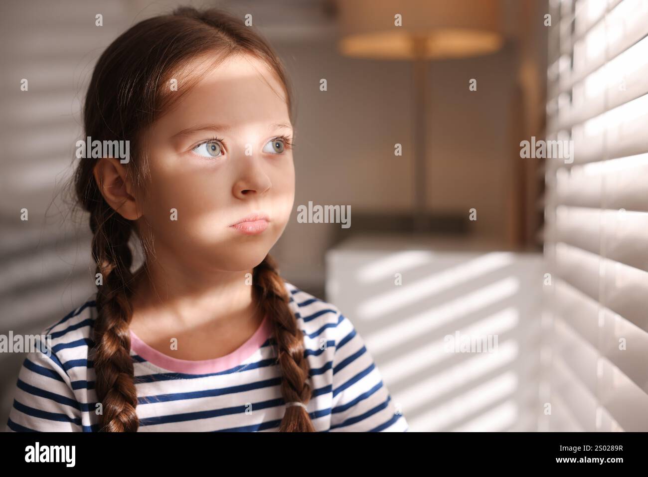Orphanage concept. Sad girl and looking through window blinds indoors ...