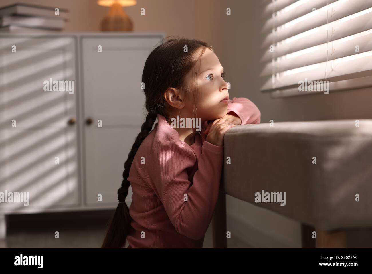 Orphanage concept. Sad girl and looking through window blinds indoors ...